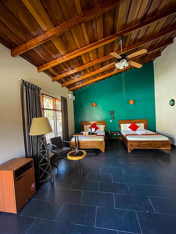 Quarto de hotel com duas camas de madeira, um ventilador de teto, uma escrivaninha com cadeira e uma janela com cortinas.