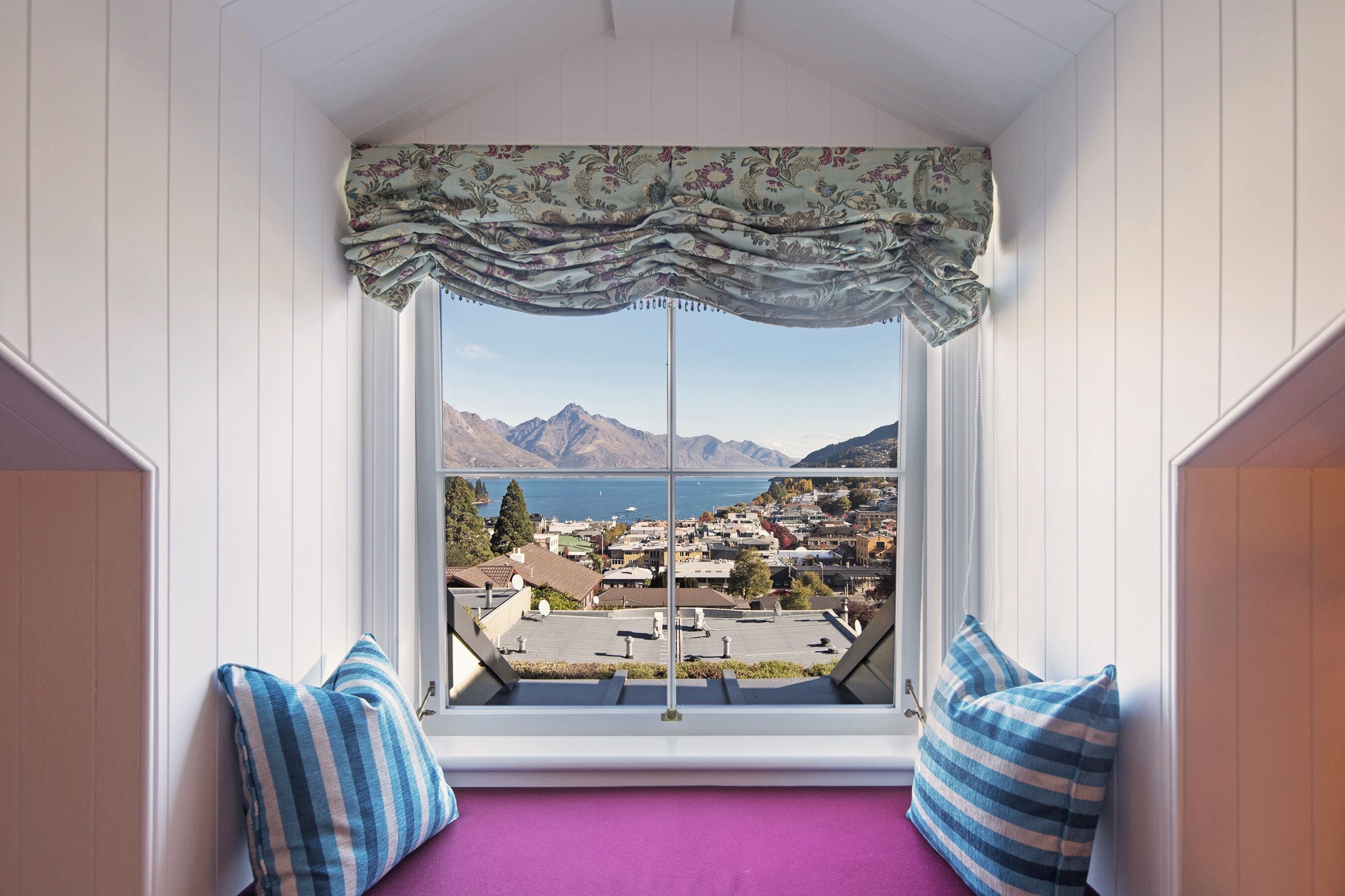 Quarto com uma grande janela com vista para uma cidade e montanhas. Há dois travesseiros no peitoril da janela.