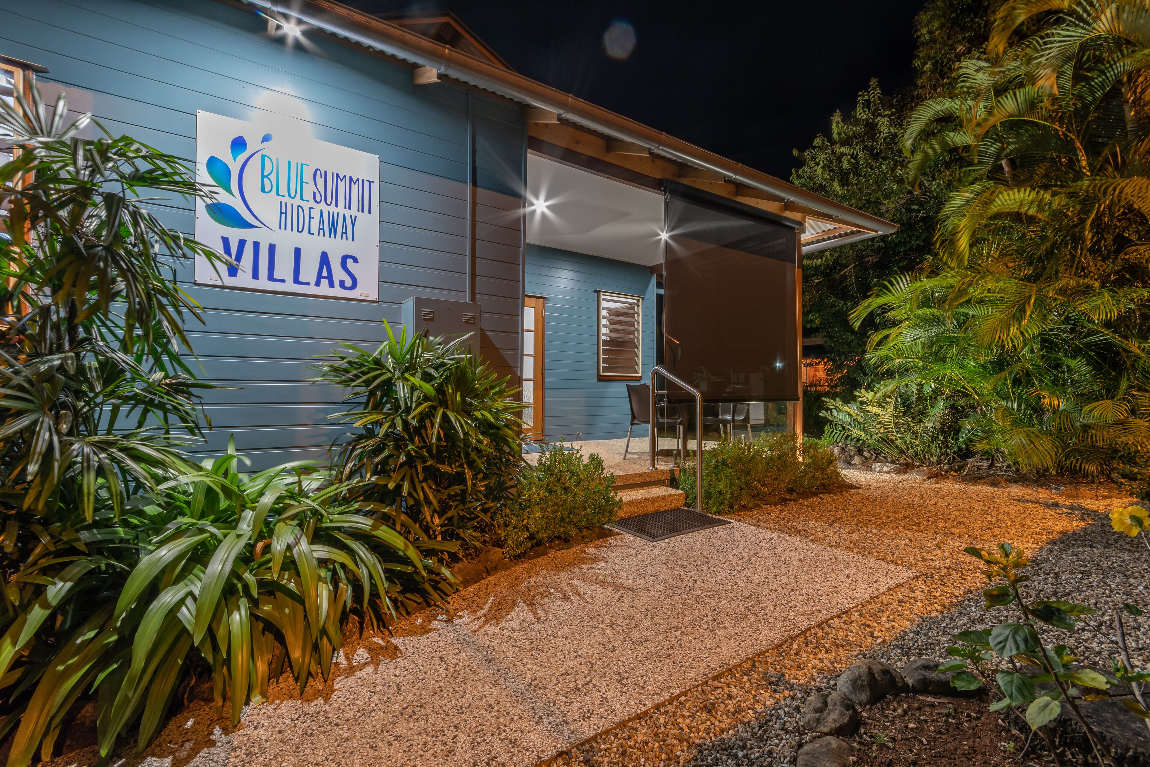 Um hotel chamado 'Bluesummit Hideaway Villas' com uma entrada bem iluminada, um caminho de cascalho e vegetação exuberante ao redor.