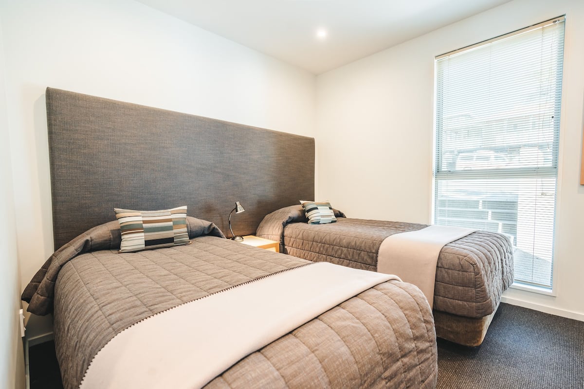 Quarto de hotel com duas camas, uma cabeceira grande, uma mesa de cabeceira com luminária e uma janela com persianas.