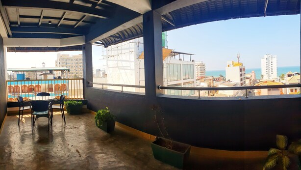 Varanda com vista para a cidade e o mar, com mesa, cadeiras, plantas em vasos e grade de metal.