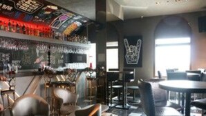 Uma área de bar com diversos tipos de garrafas e copos, banquetas e um cartaz grande na parede.