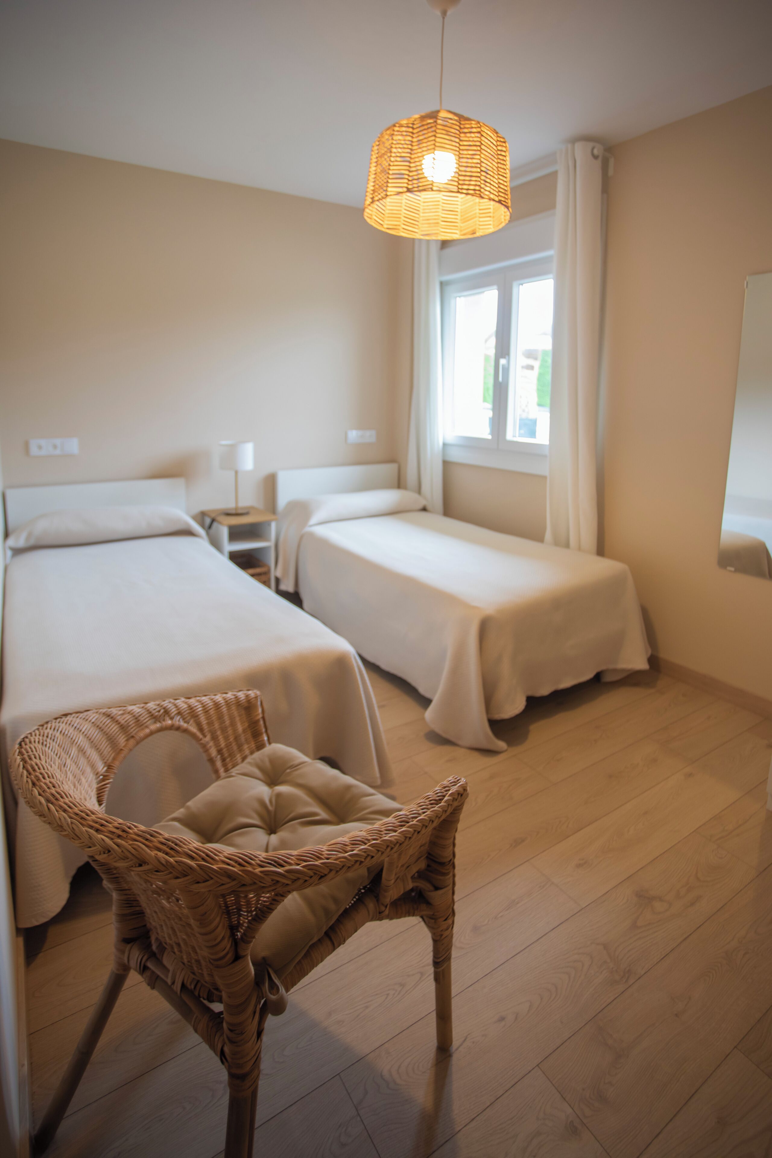 Quarto de hotel com duas camas, uma cadeira de vime, uma mesa de cabeceira com abajur e uma janela com cortinas.