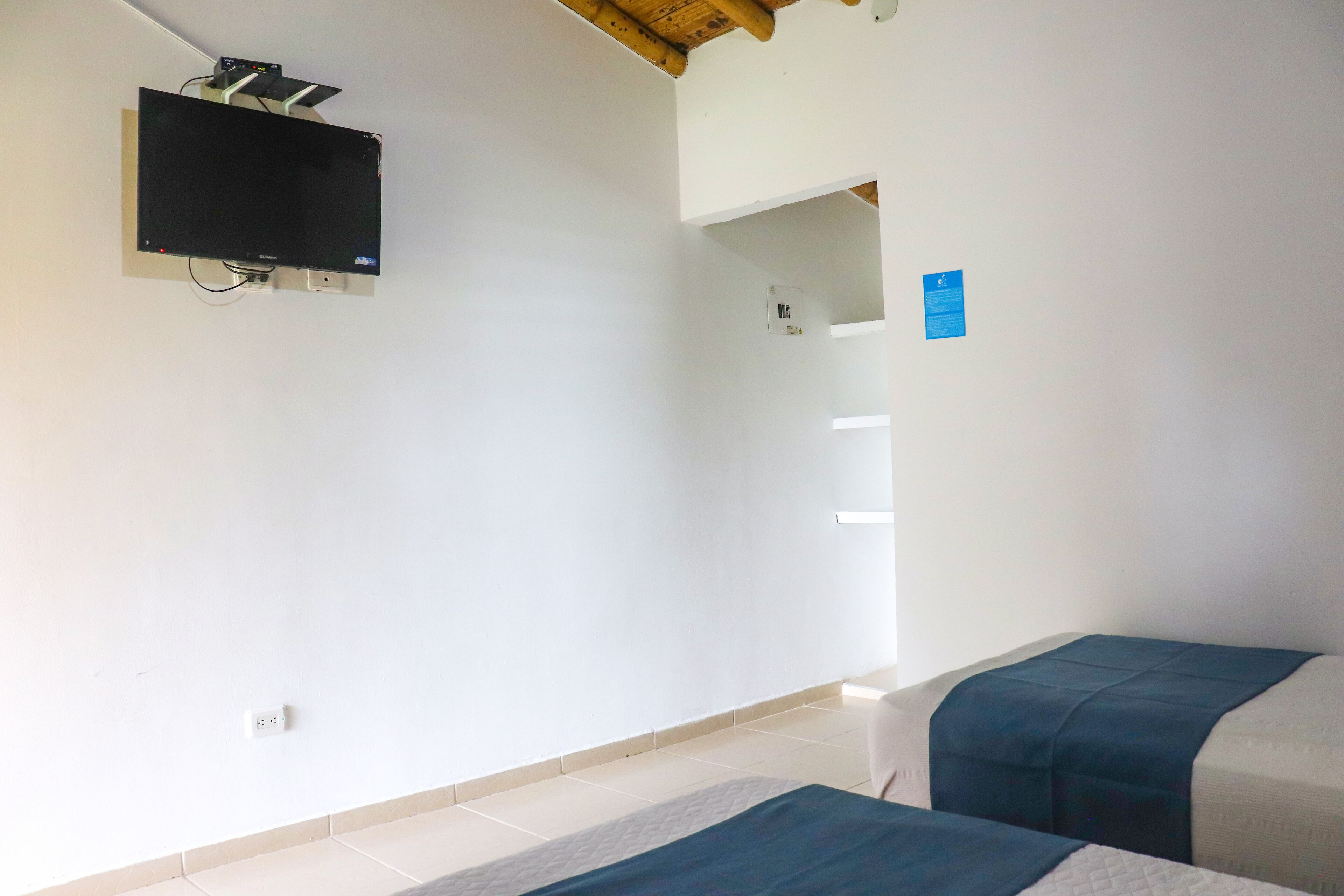 Quarto com televisão de tela plana na parede, cama com manta azul e parede branca com uma pequena placa azul.