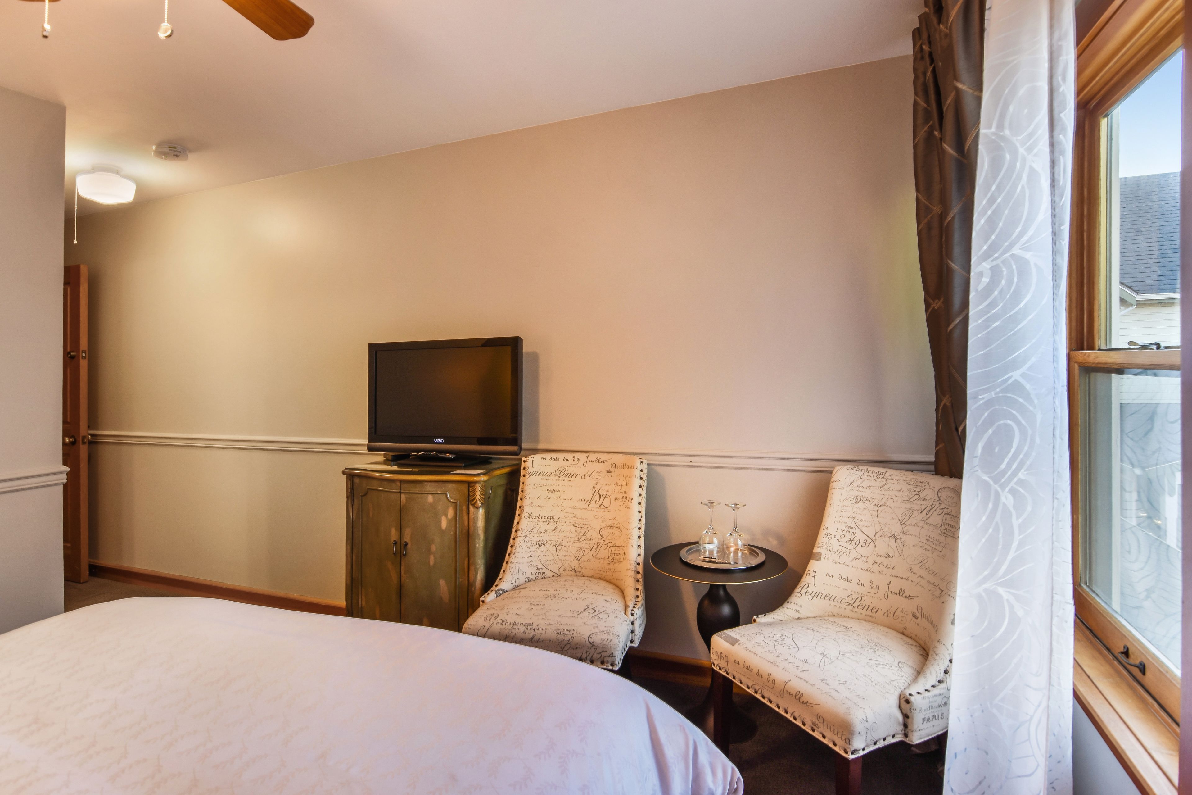 Quarto de hotel com uma cama, uma televisão sobre um móvel de madeira, duas poltronas estofadas com padrões decorativos, uma mesinha com dois copos de vinho e uma janela com cortinas.