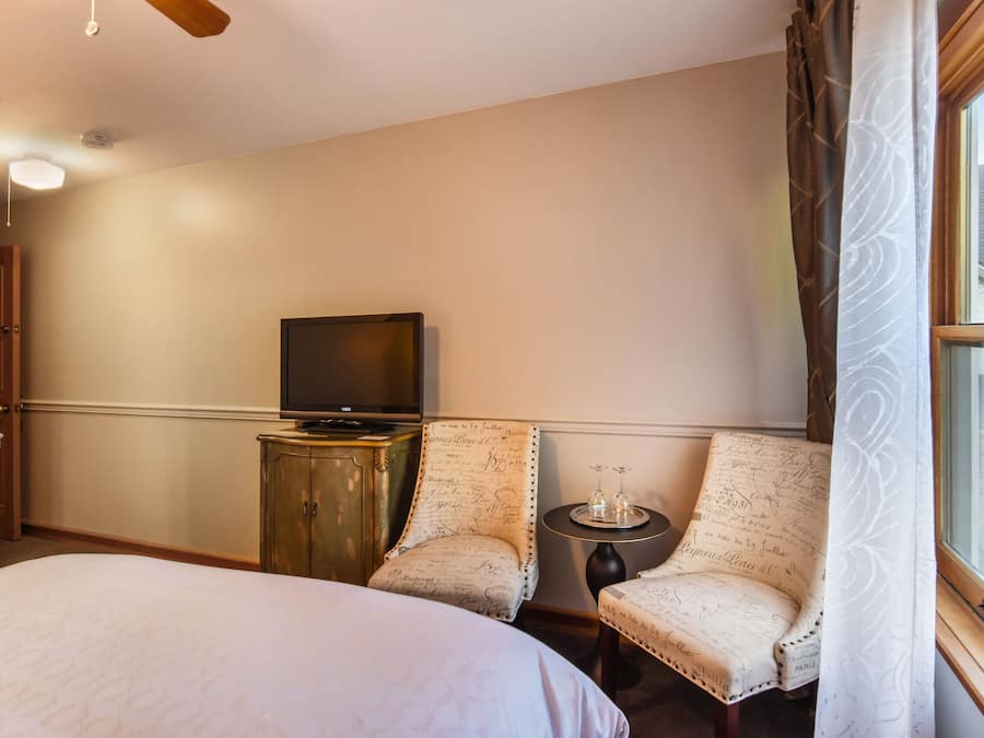 Quarto de hotel com uma cama, uma televisão sobre um móvel de madeira, duas poltronas estofadas com padrões decorativos, uma mesinha com dois copos de vinho e uma janela com cortinas.