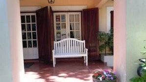 Uma área externa coberta com um banco branco, um vaso com flores e uma porta com cortinas.