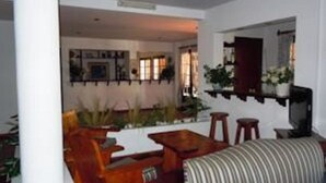Uma sala de estar com um sofá, uma mesa de centro e uma televisão. Há uma planta e uma estante com objetos decorativos.