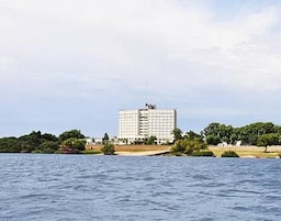 Um edifício branco e imponente com vários andares, localizado perto de um corpo de água e rodeado por vegetação.