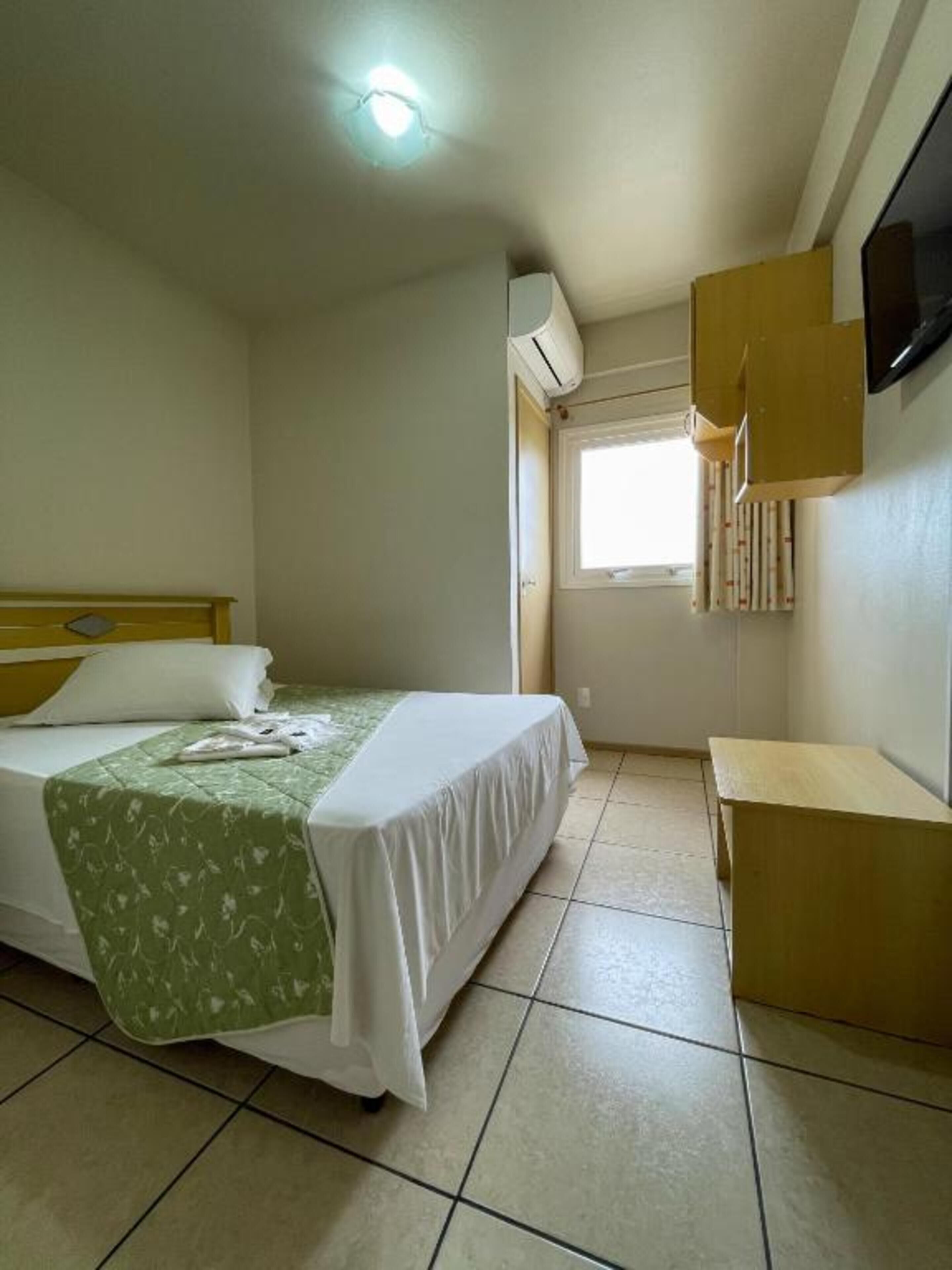 Quarto de hotel com cama, uma pequena mesa de madeira, televisão fixada na parede e aparelho de ar condicionado.