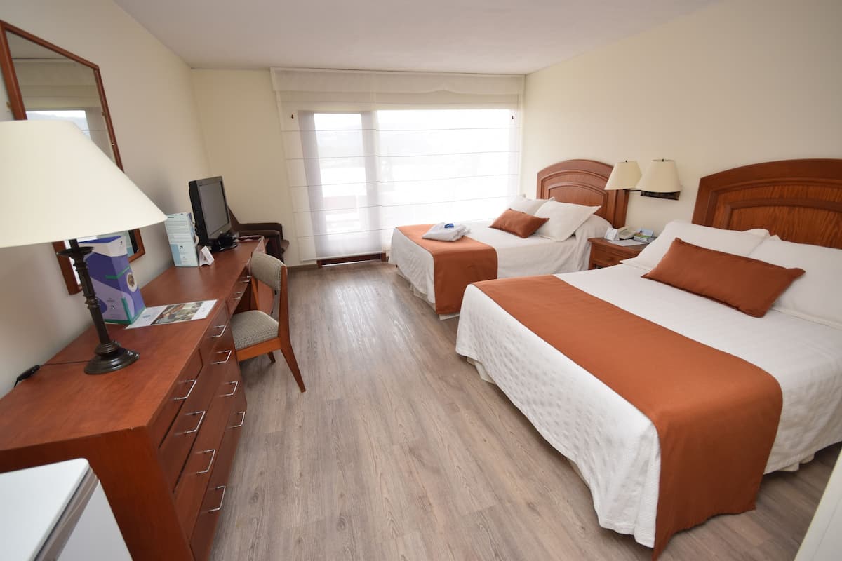Quarto de hotel com duas camas, uma escrivaninha com computador, uma cadeira e uma janela com cortinas.