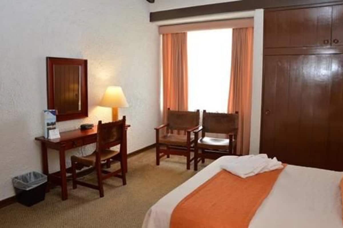 Quarto de hotel com uma cama, uma escrivaninha com luminária, duas cadeiras e uma janela com cortinas.