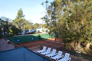 Uma quadra de tênis com cerca verde, cadeiras de lounge brancas e árvores ao fundo.