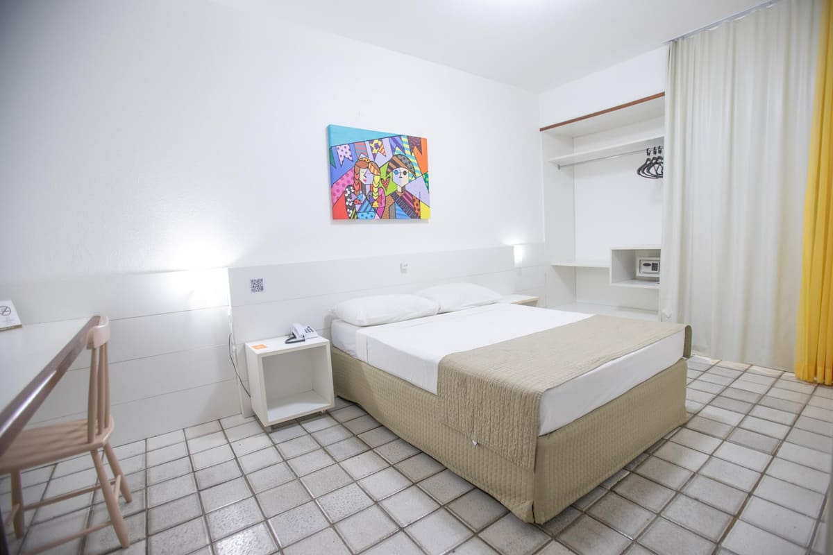 Quarto de hotel com uma cama, uma cadeira, uma mesinha de cabeceira, um quadro na parede e banheiro com cortina de chuveiro.