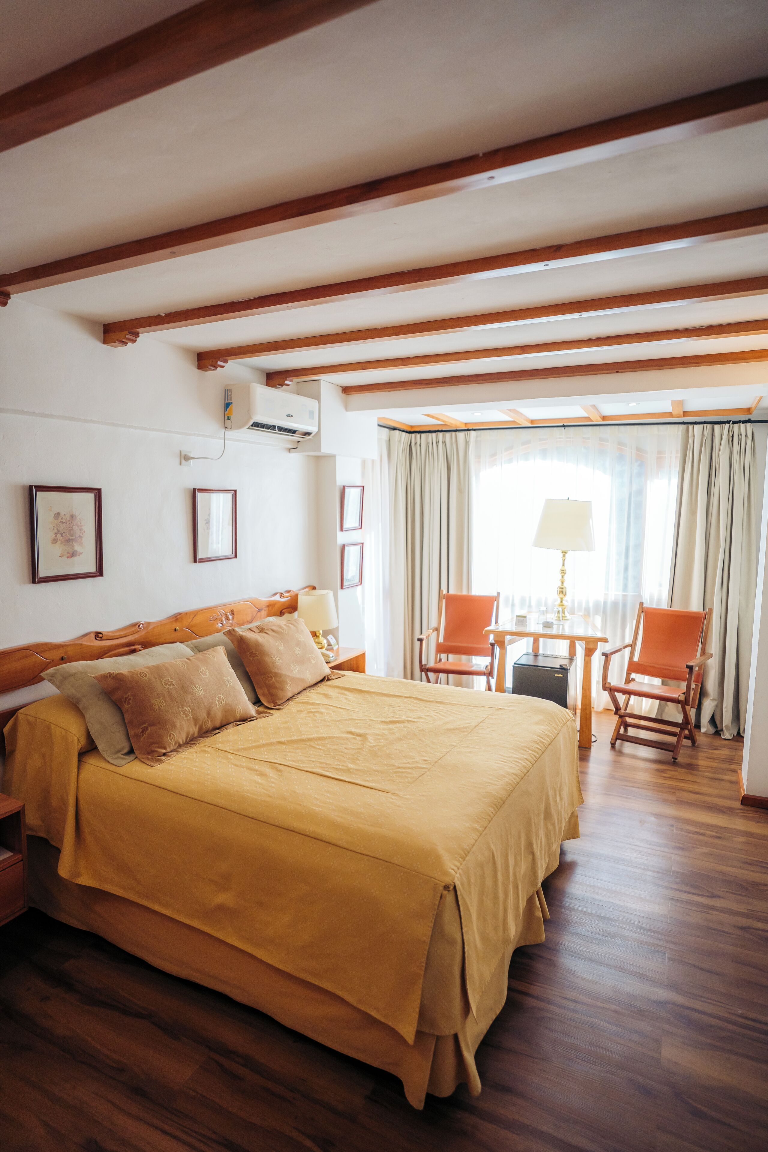 Quarto de hotel com uma cama grande, vigas de madeira, uma pequena mesa com cadeiras e uma janela com cortinas.