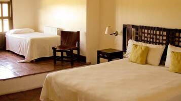 Quarto de hotel com duas camas individuais, uma cadeira de madeira, uma mesinha de cabeceira e uma luminária na parede.