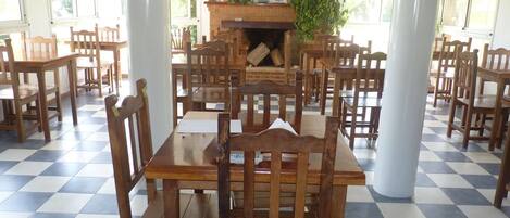 Uma sala de jantar com mesas e cadeiras de madeira, chão xadrez e lareira com uma planta em cima.