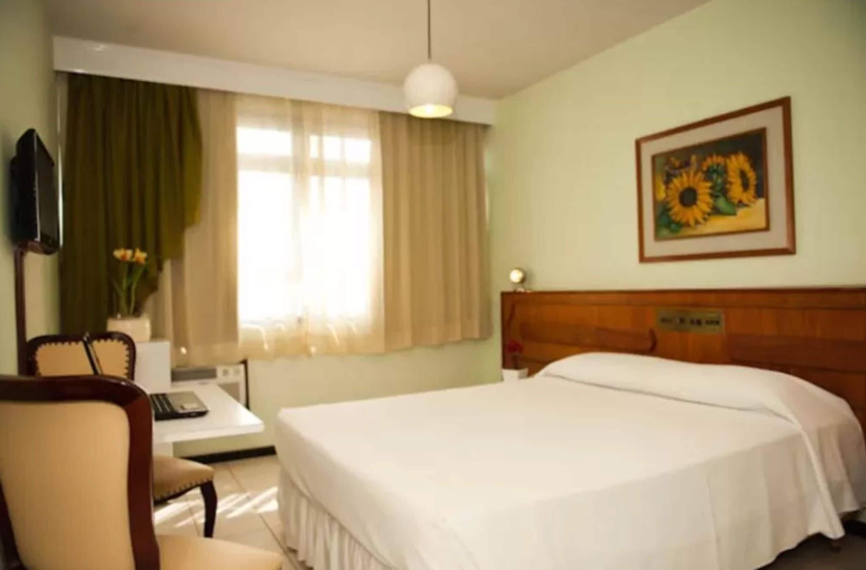 Quarto de hotel com cama, escrivaninha com cadeira, televisão e janela com cortinas.