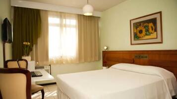 Quarto de hotel com cama, escrivaninha com cadeira, televisão e janela com cortinas.