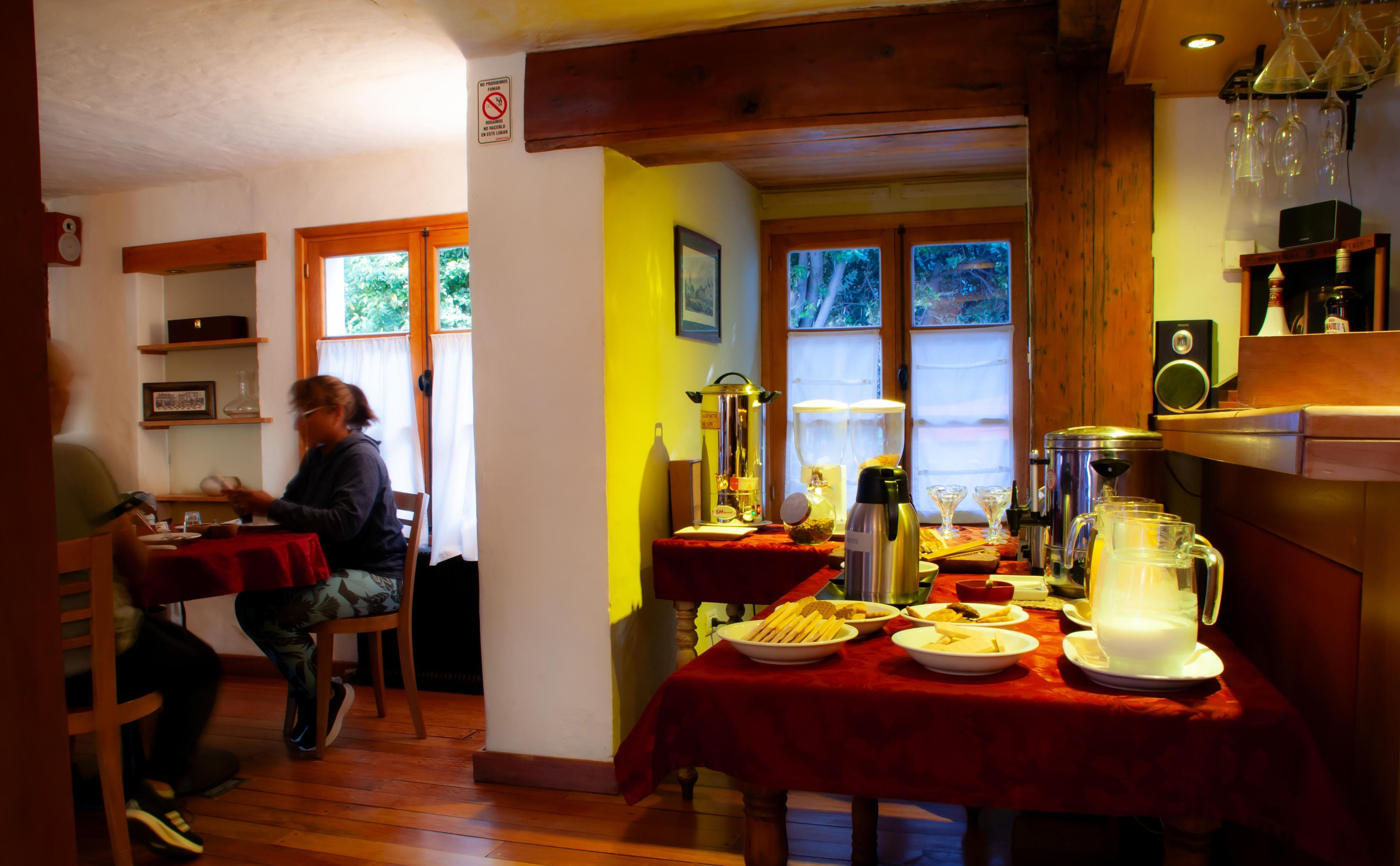 Uma área de refeições com uma mesa posta para dois, cafeteira e janela com cortinas.