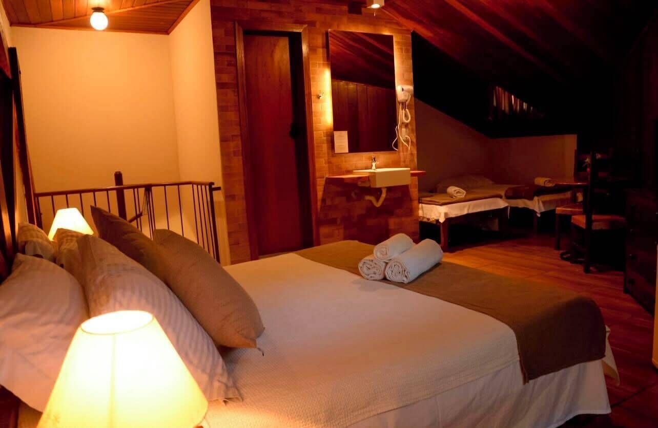 Uma cama de solteiro com almofadas e uma toalha enrolada, uma mesa de cabeceira, um espelho e um banheiro pequeno com pia e toalha.
