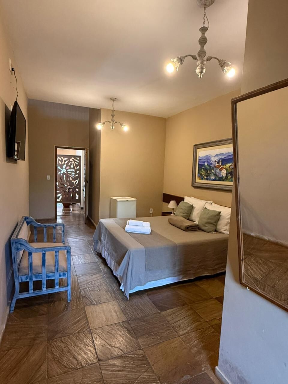 Quarto de hotel com uma cama, uma cadeira, uma escrivaninha, um candelabro e um quadro na parede.
