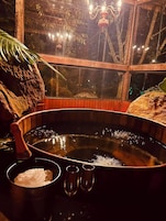 Uma banheira de hidromassagem com estrutura de madeira, uma janela com vista para árvores e um candelabro.