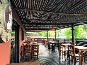 Um pátio ao ar livre com mesas e cadeiras de madeira, teto de bambu e vista para o verde ao fundo.