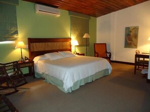 Quarto de hotel com uma cama grande, duas mesas de cabeceira com abajures, uma cadeira de balanço de madeira e uma escrivaninha com cadeira.