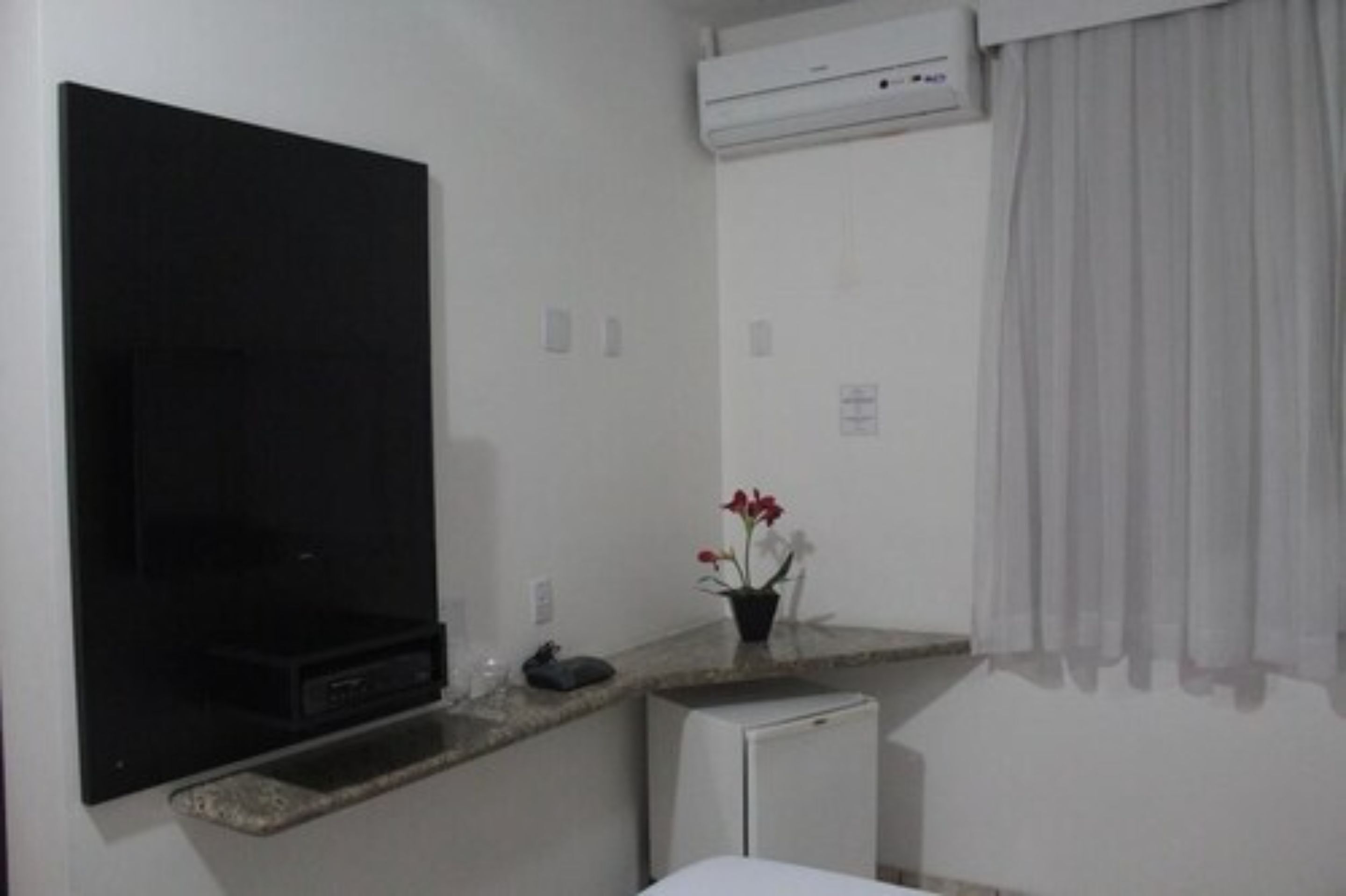 Quarto pequeno com televisão de tela plana na parede, uma geladeira compacta, um vaso de planta e uma janela com cortinas.