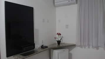 Quarto pequeno com televisão de tela plana na parede, uma geladeira compacta, um vaso de planta e uma janela com cortinas.