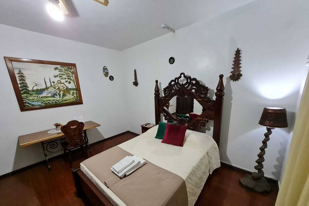 Quarto com cama de madeira, escrivaninha e cadeira. Há uma pintura na parede e um abajur no chão.