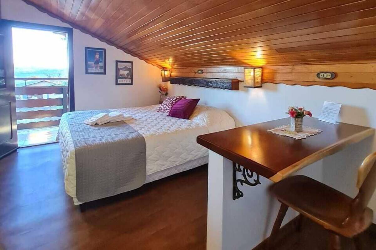 Um quarto aconchegante com teto de madeira, cama com colcha branca e cinza, uma mesinha com um vaso de flores e uma cadeira.