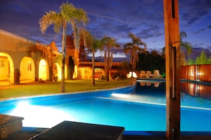 Uma piscina com espreguiçadeiras, palmeiras e um edifício com arcos ao fundo.