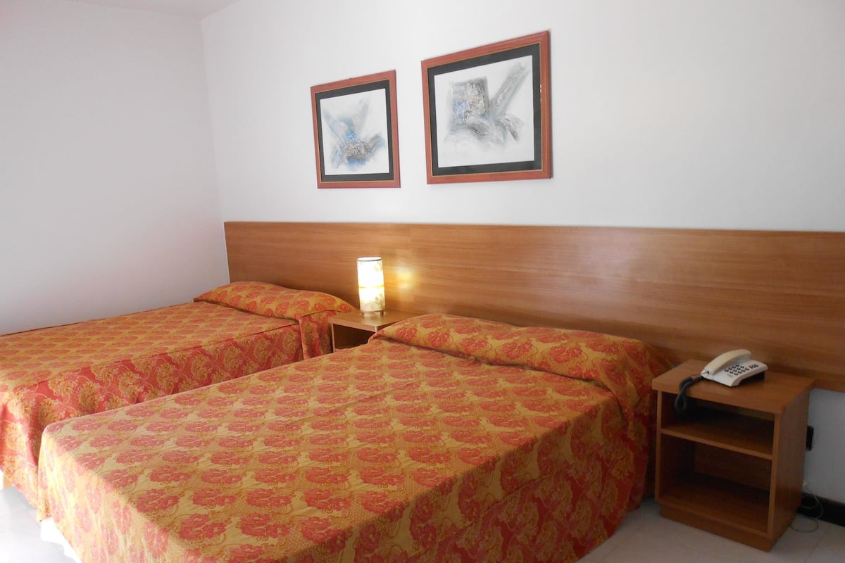 Quarto de hotel com duas camas de solteiro, uma mesa de cabeceira com abajur, telefone e duas fotos emolduradas na parede.