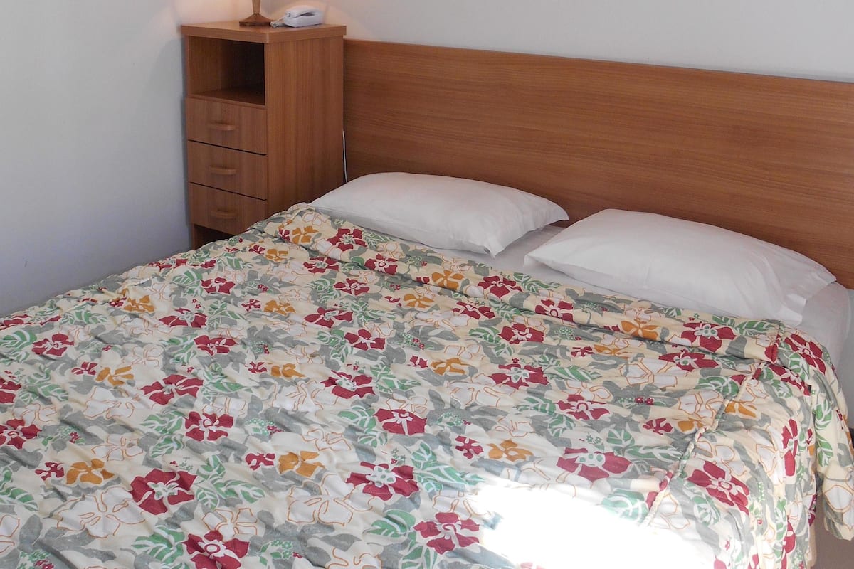 Uma cama bem arrumada com colcha florida, luminária de cabeceira, mesa de cabeceira de madeira com um telefone e um quadro na parede.