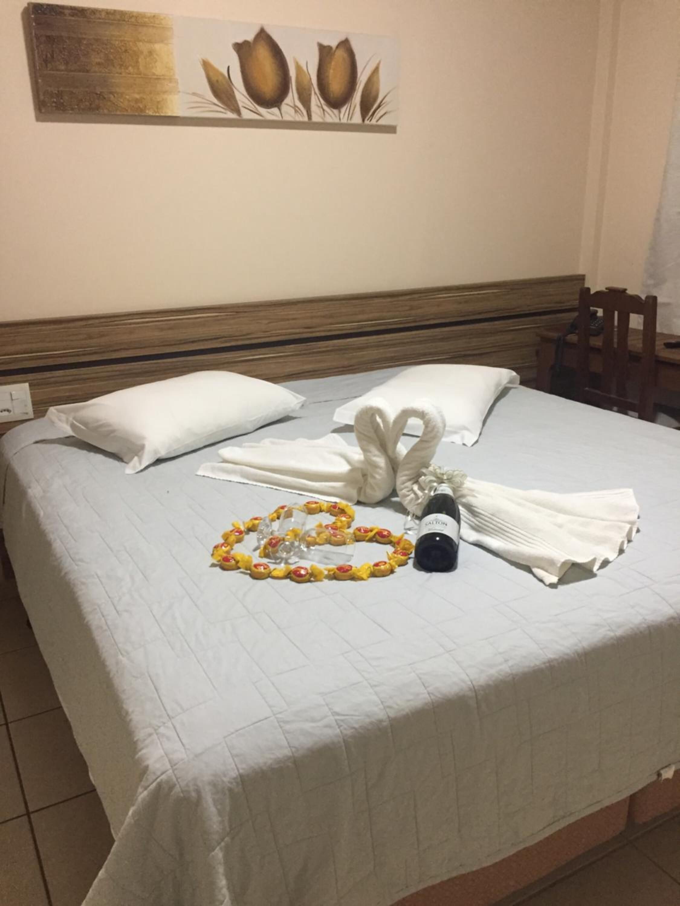 Uma cama bem arrumada com roupa de cama branca, uma toalha em formato de coração, uma garrafa de água e uma guirlanda de flores amarelas.