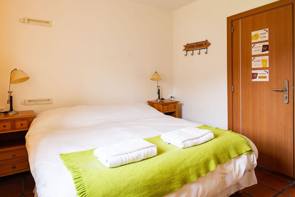 Quarto de hotel com cama, duas mesas de cabeceira, luminária, radiador, porta de madeira e placa na parede com comodidades.