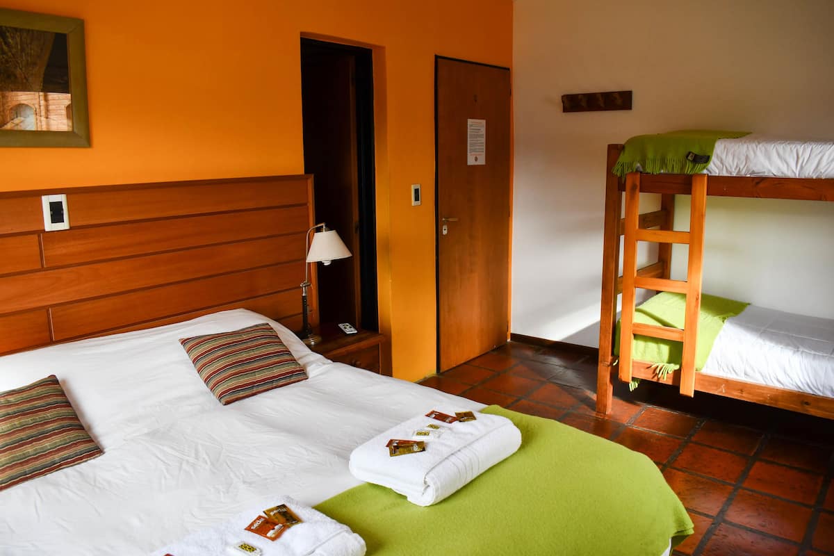 Quarto de hotel com uma cama grande, uma cama menor em beliche, mesa de cabeceira com abajur e cabeceira de madeira.