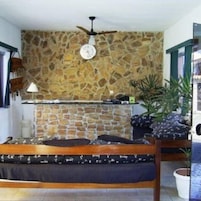 Quarto com parede de pedra, cama com cabeceira escura, ventilador de teto e um relógio.