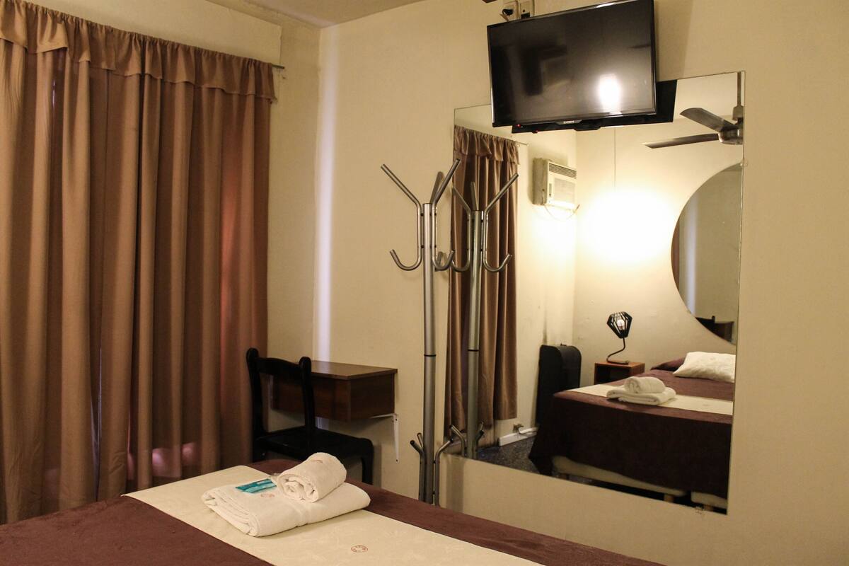Quarto de hotel com cama, escrivaninha, cadeira, televisão, espelho e porta-roupas.