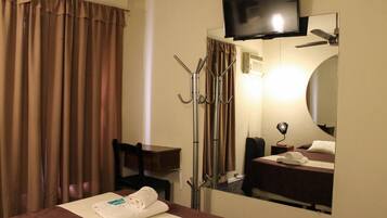 Quarto de hotel com cama, escrivaninha, cadeira, televisão, espelho e porta-roupas.