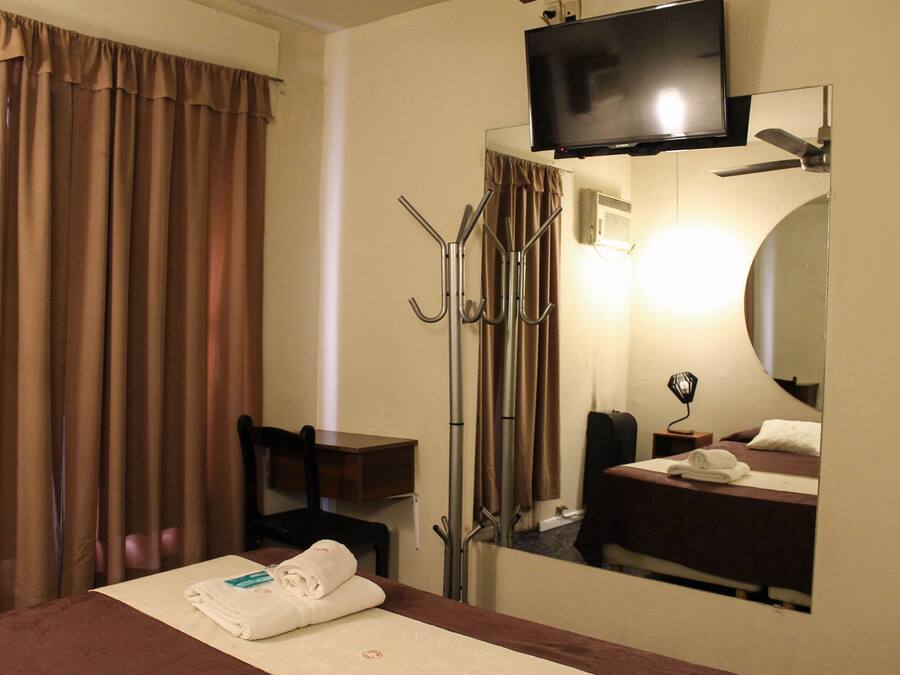 Quarto de hotel com cama, escrivaninha, cadeira, televisão, espelho e porta-roupas.