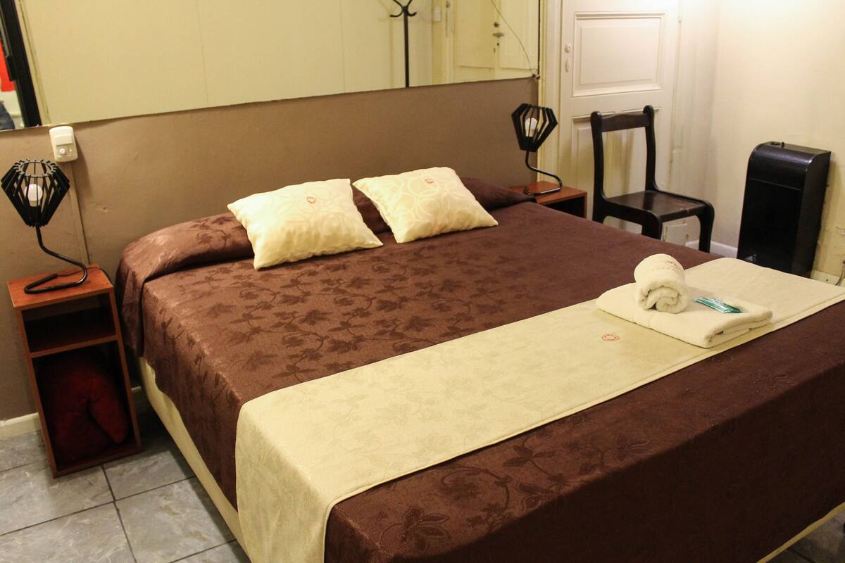 Uma cama bem arrumada com um cobre-leito marrom e duas almofadas, uma mesa de cabeceira com abajur, uma cadeira e um alto-falante.