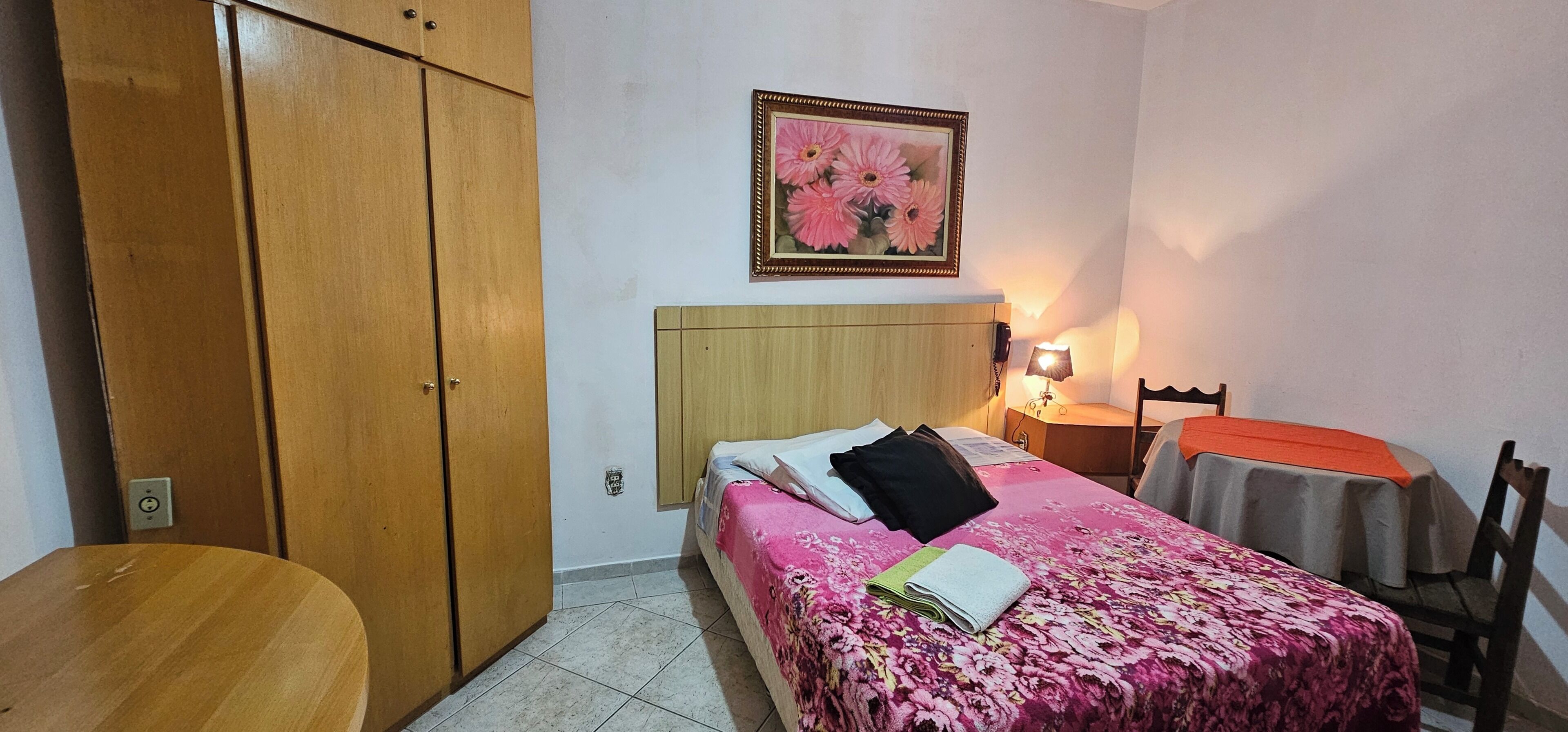 Quarto com cama, guarda-roupa de madeira, mesa, cadeira, abajur e quadro na parede.