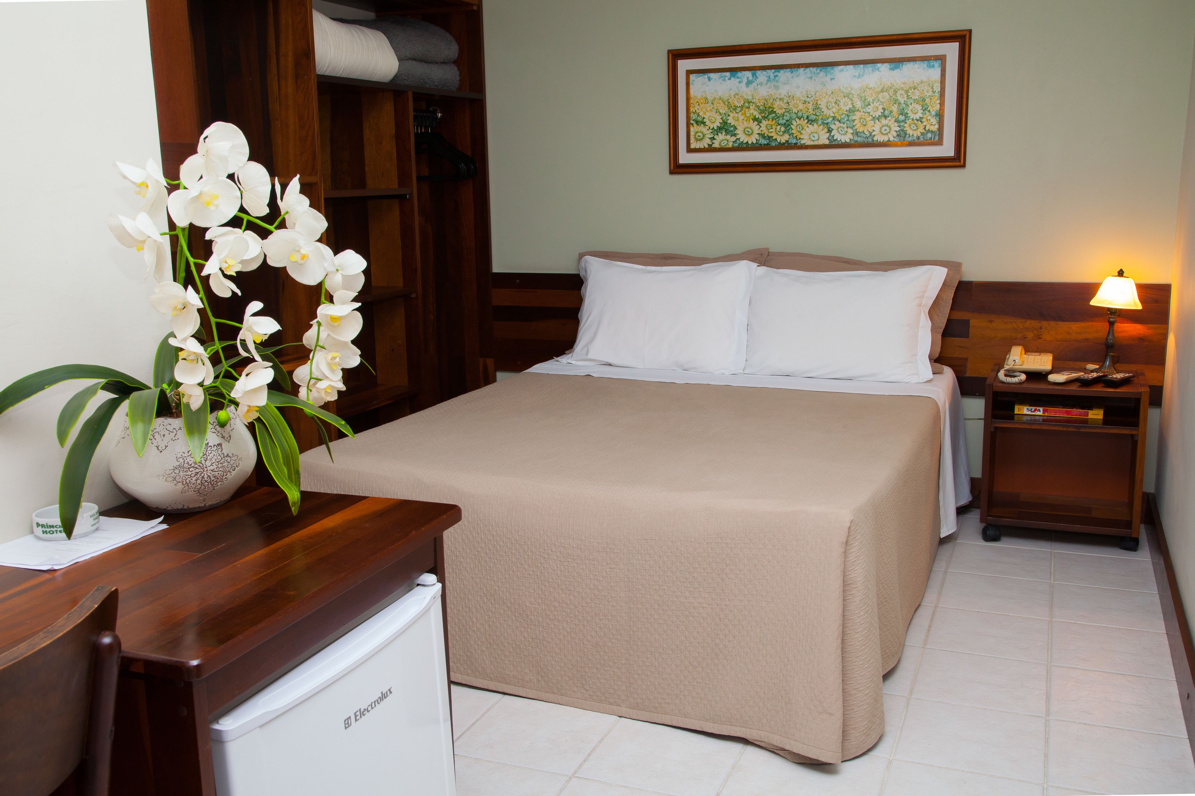Cama bem arrumada com colcha bege, luminária de cabeceira, mesa de cabeceira com telefone e um vaso de orquídeas brancas sobre uma escrivaninha de madeira.