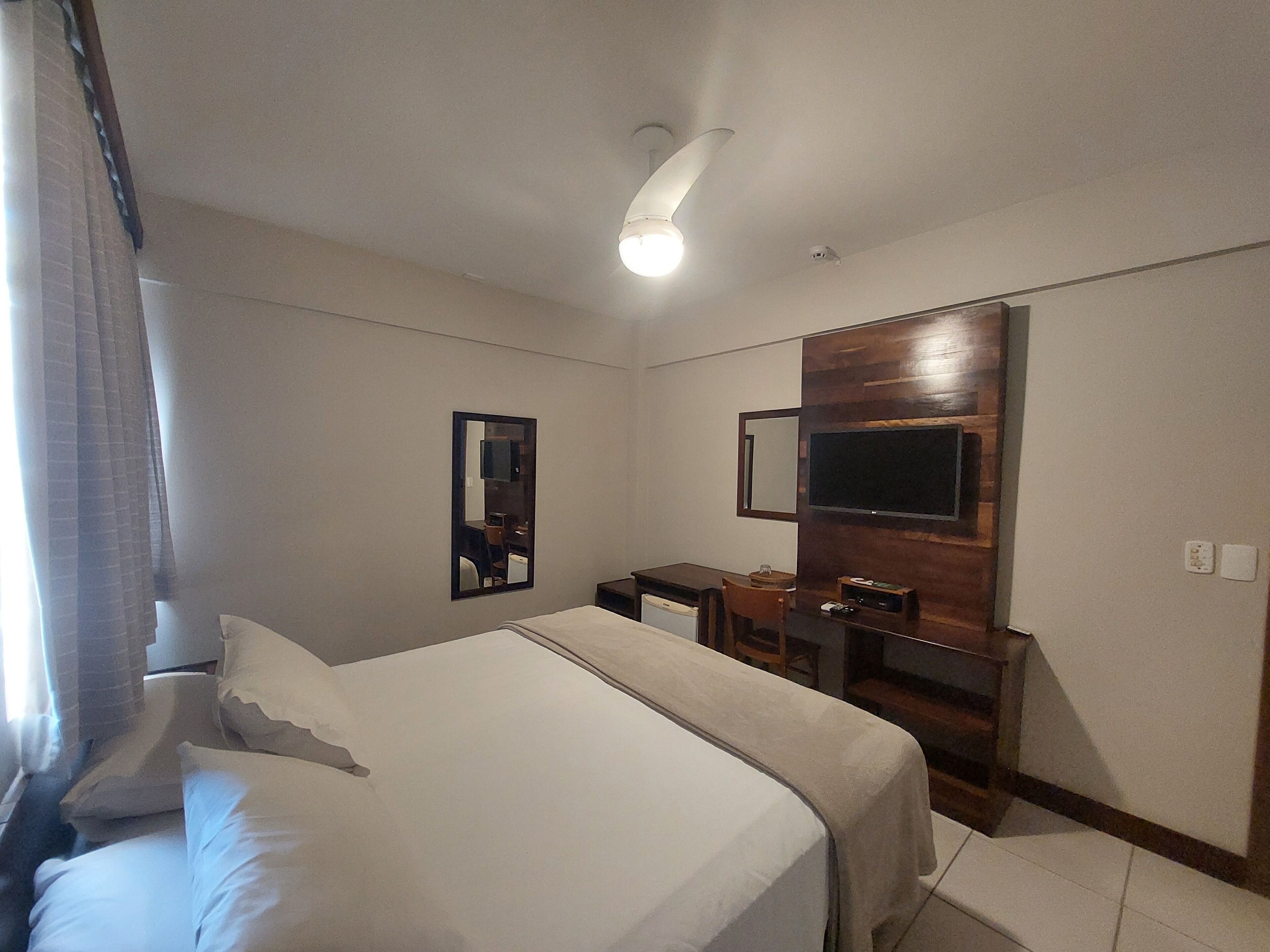 Quarto de hotel com cama, escrivaninha, cadeira, espelho e televisão.