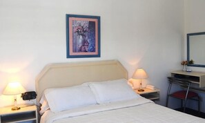 Quarto de hotel com cama, duas luminárias de cabeceira, mesa de apoio, escrivaninha com cadeira, espelho e uma obra de arte emoldurada na parede.