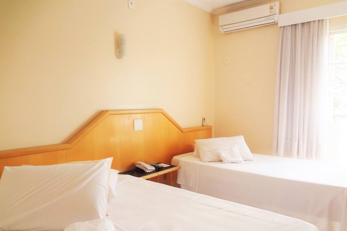 Quarto de hotel com duas camas de solteiro, cabeceira de madeira, mesa de cabeceira com telefone e ar condicionado.