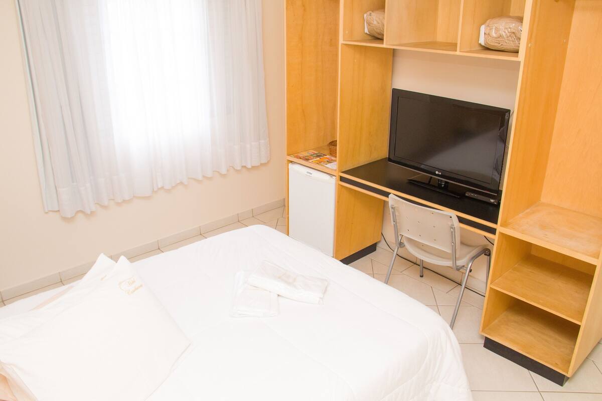 Quarto de hotel com cama, escrivaninha com televisão, cadeira e um pequeno frigorífico.